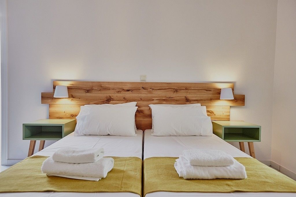 Uma cama de casal com roupa de cama branca e cabeceira de madeira, com duas mesas de cabeceira e abajures de cada lado.