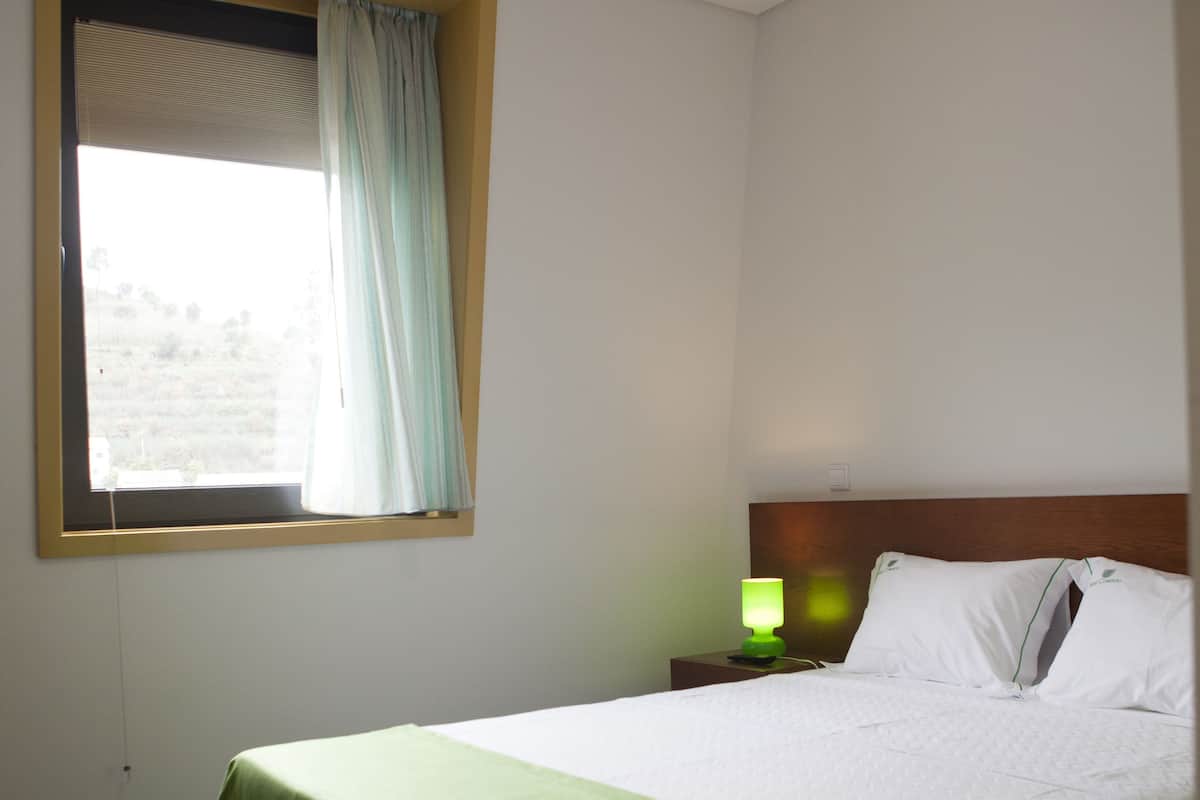 Quarto de hotel com uma cama de solteiro, mesa de cabeceira com abajur verde, janela com cortina e vista para o exterior.