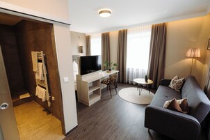 Quarto de hotel moderno com televisão de ecrã plano, um sofá, uma pequena mesa de jantar e uma planta.