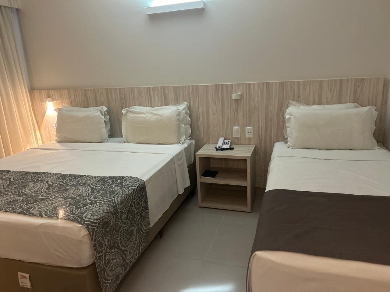 Quarto de hotel com duas camas, uma mesinha de cabeceira com telefone e uma luminária na parede.