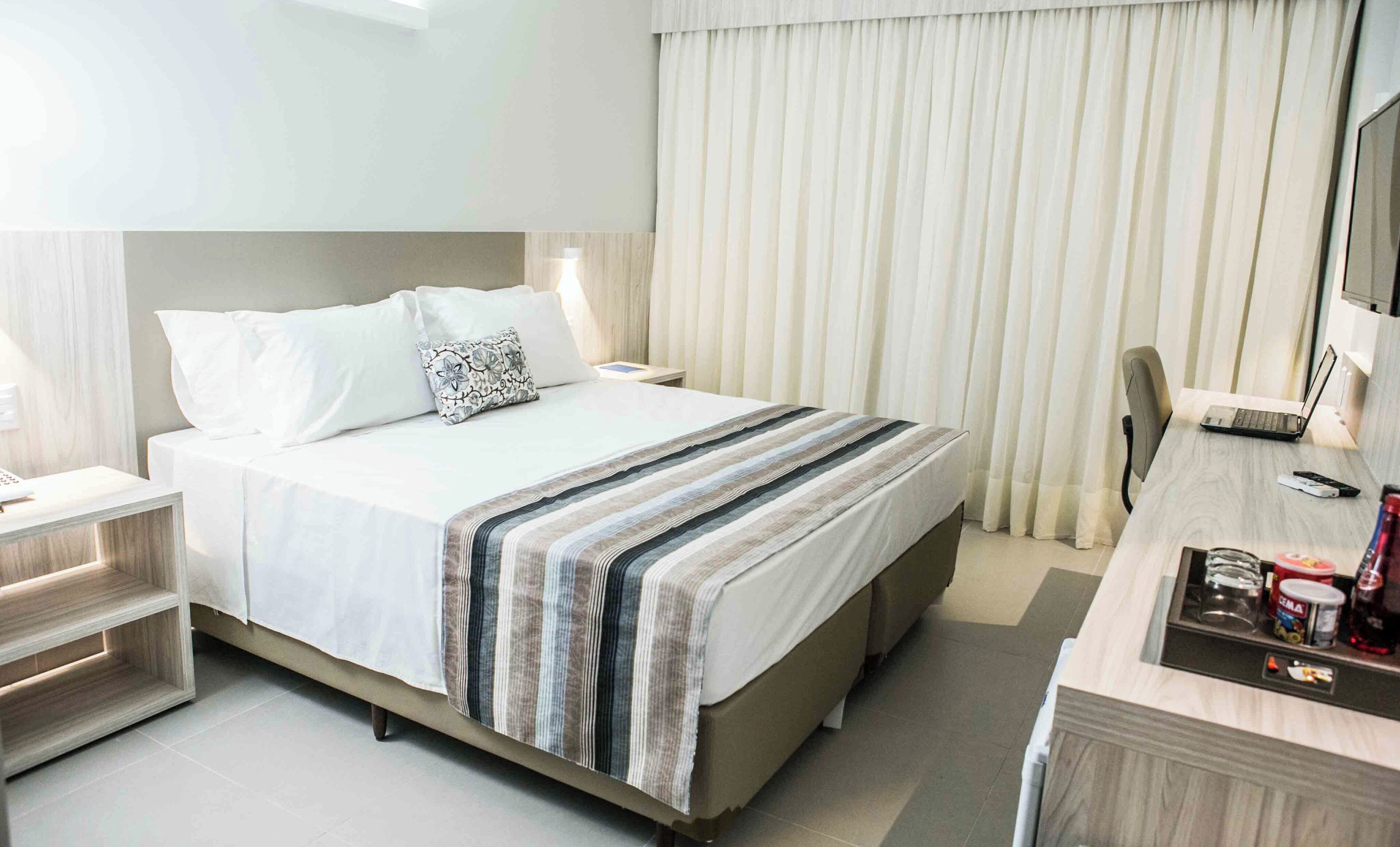 Quarto de hotel com cama, mesa de cabeceira e escrivaninha com um laptop.