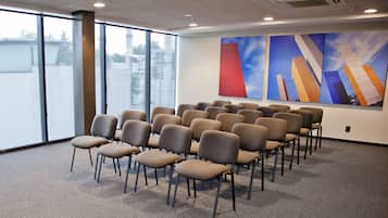 Uma sala de conferências com fileiras de cadeiras, janelas amplas e um mural de arte moderna na parede.