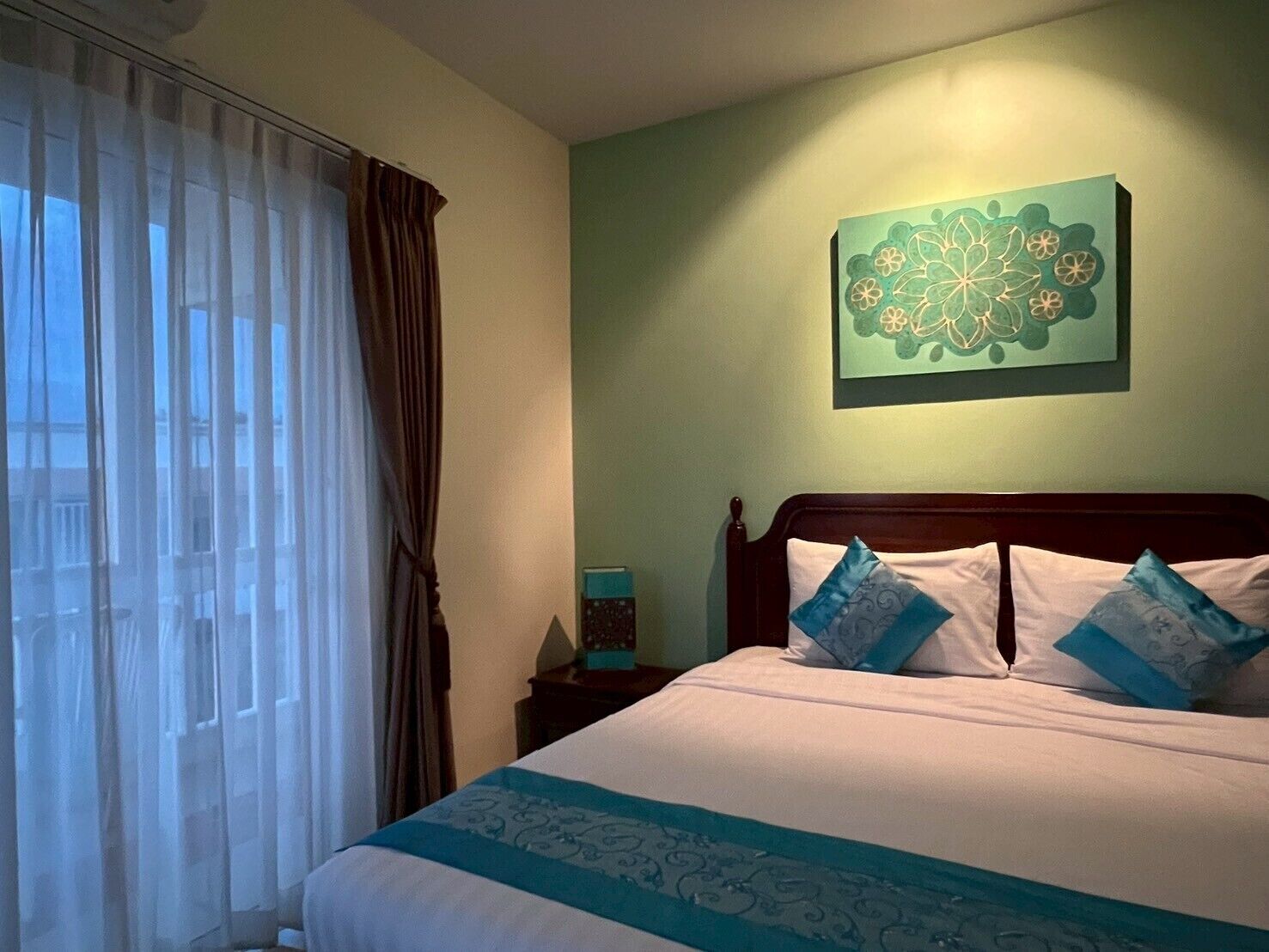 Uma cama bem arrumada com colcha azul e branca, um objeto decorativo na parede acima dela, e uma janela com cortinas leves.