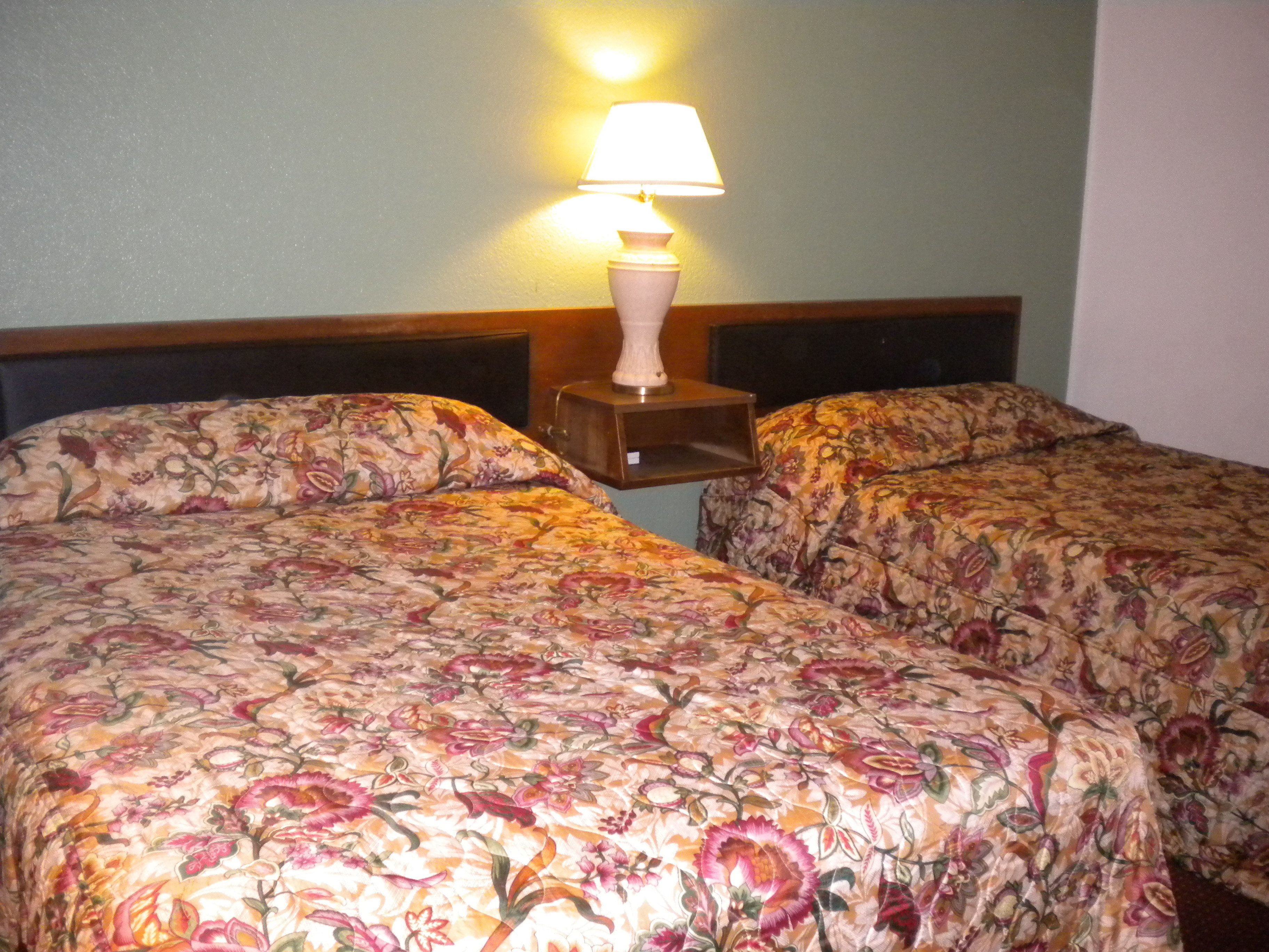 Quarto de hotel com duas camas, cada uma com roupa de cama estampada com motivos florais, uma mesa de cabeceira com luminária e cabeceira de madeira.