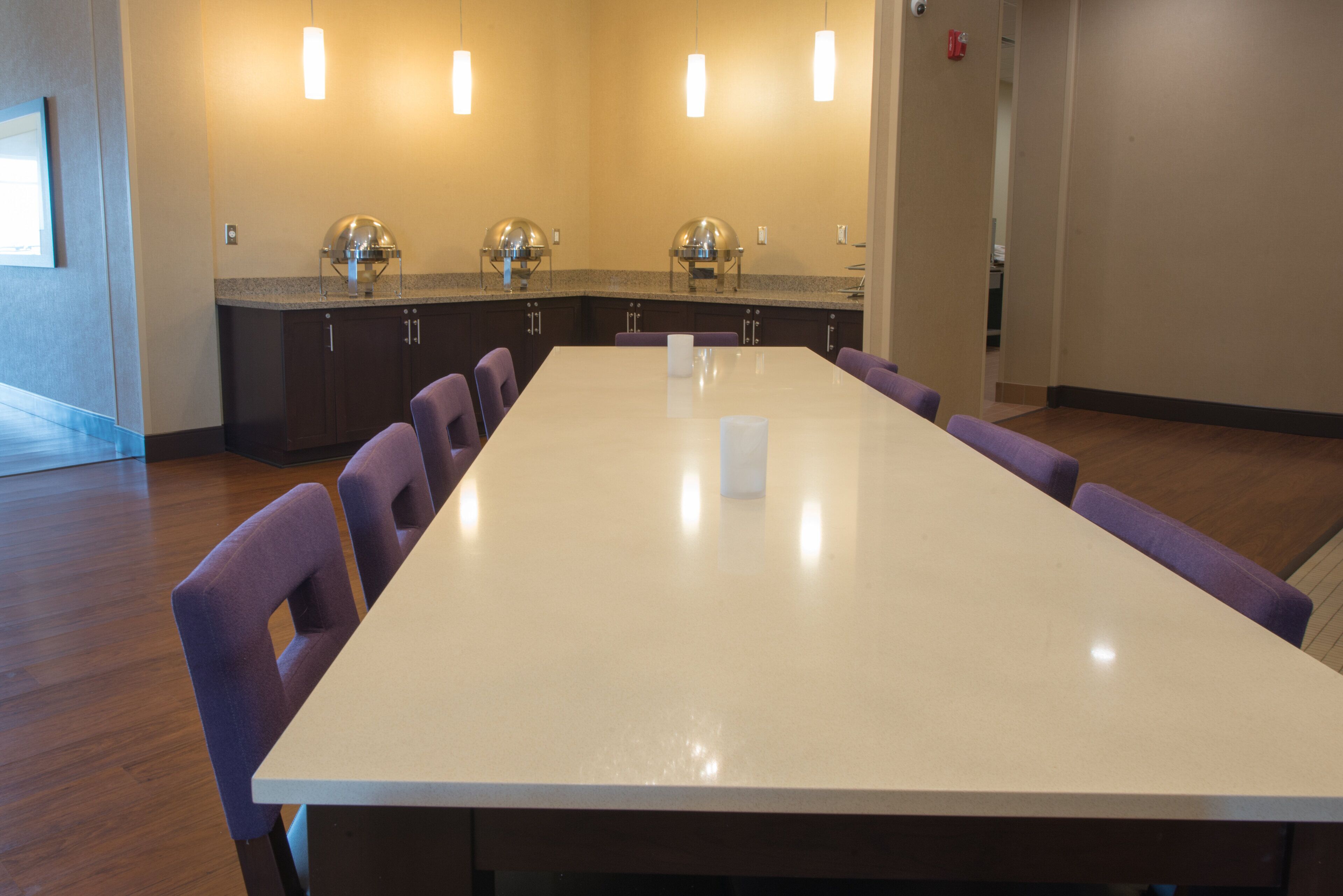 Uma sala de conferência com uma mesa comprida, cadeiras roxas e uma área de cozinha com bancada de granito e eletrodomésticos de aço inoxidável.