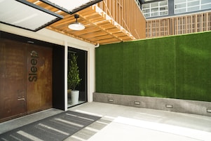 Entrada de um edifício moderno com porta de madeira com a inscrição "Sleep", parede de grama sintética verde e chão de concreto com tapete cinza.