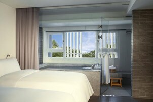 Quarto de hotel moderno com uma cama grande, chuveiro de vidro e vista para o verde lá fora, além de uma parede decorada com tijolos.