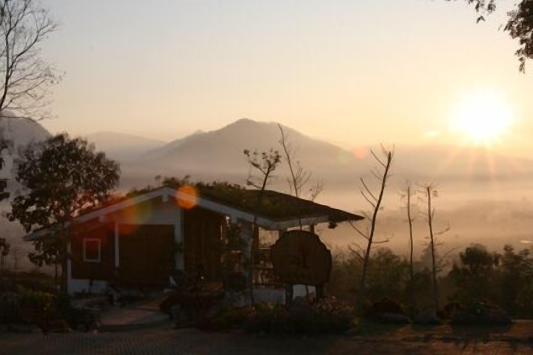 Uma cabana rústica com telhado inclinado, cercada por árvores e uma montanha ao fundo.