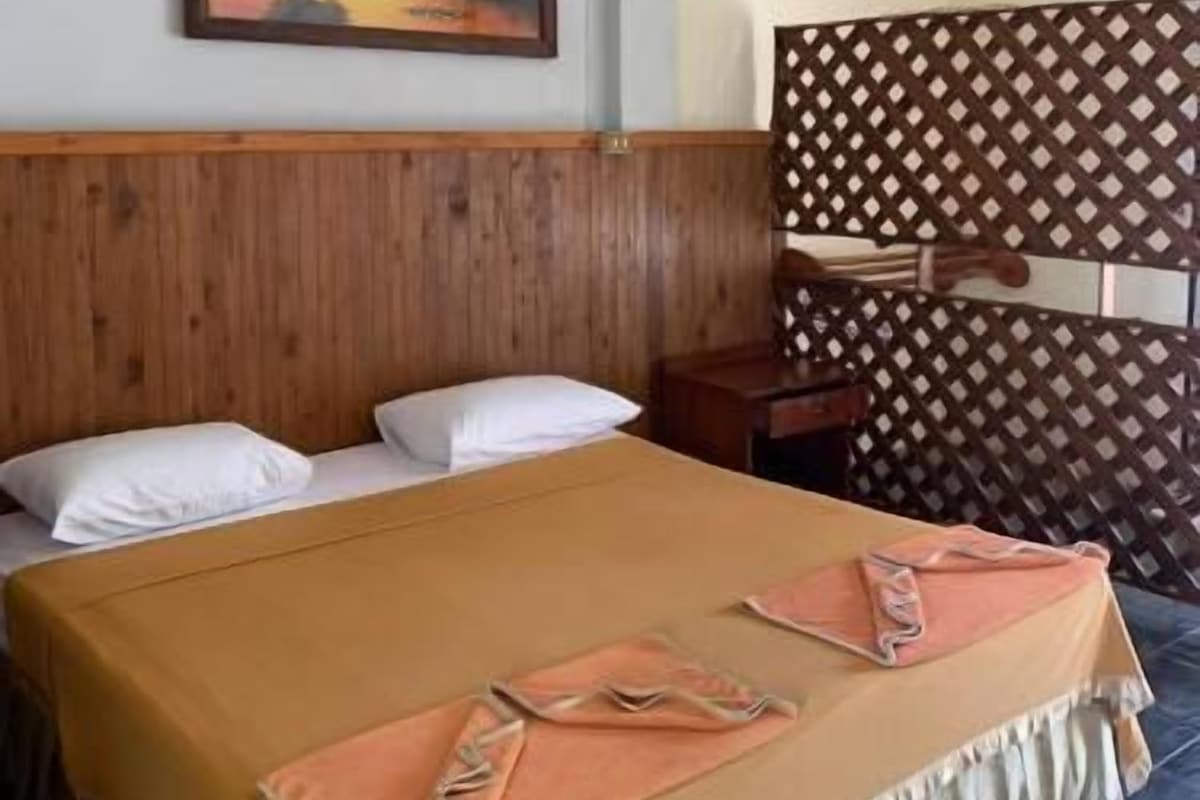 Quarto de hotel com cabeceira de madeira, cama com colcha bege, pintura na parede e divisória estampada.