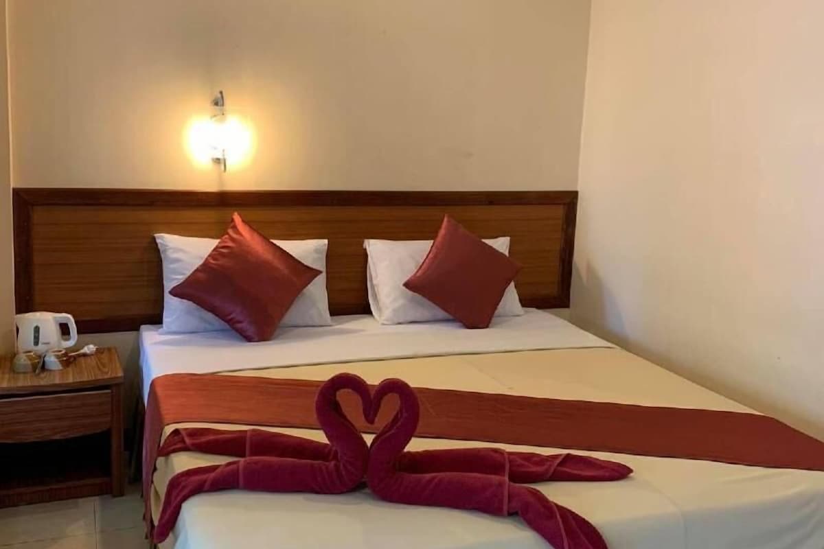 Quarto de hotel com cama, almofadas, mesa de cabeceira e luminária na parede.