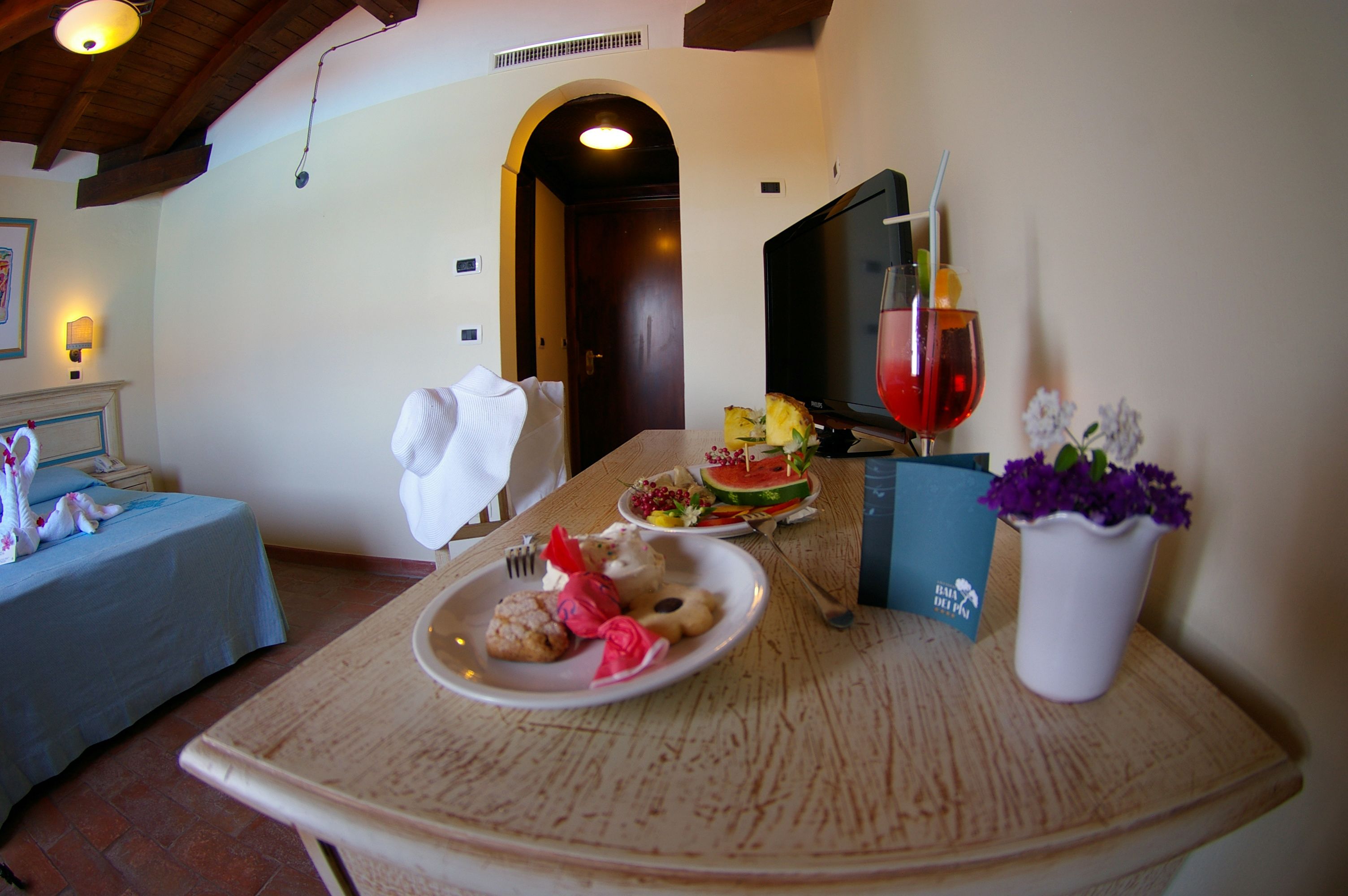 Uma sala de jantar com uma mesa de madeira, um prato de comida, um copo de bebida vermelha, uma TV e um vaso com flores.