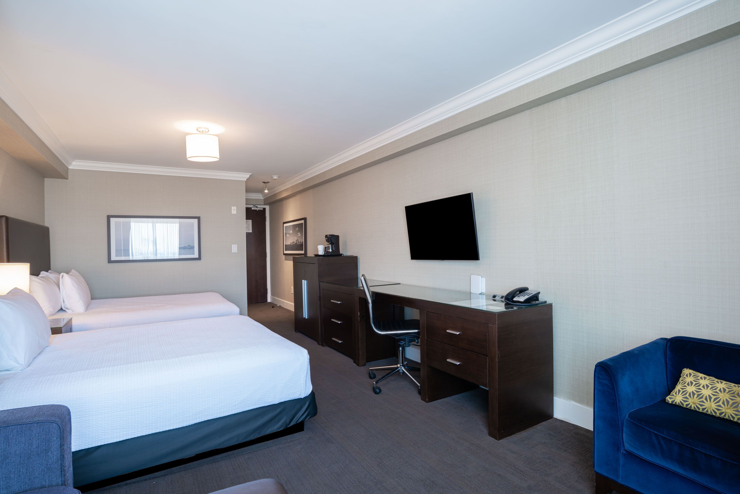 Quarto de hotel com duas camas, uma escrivaninha com cadeira, televisão e um sofá azul.