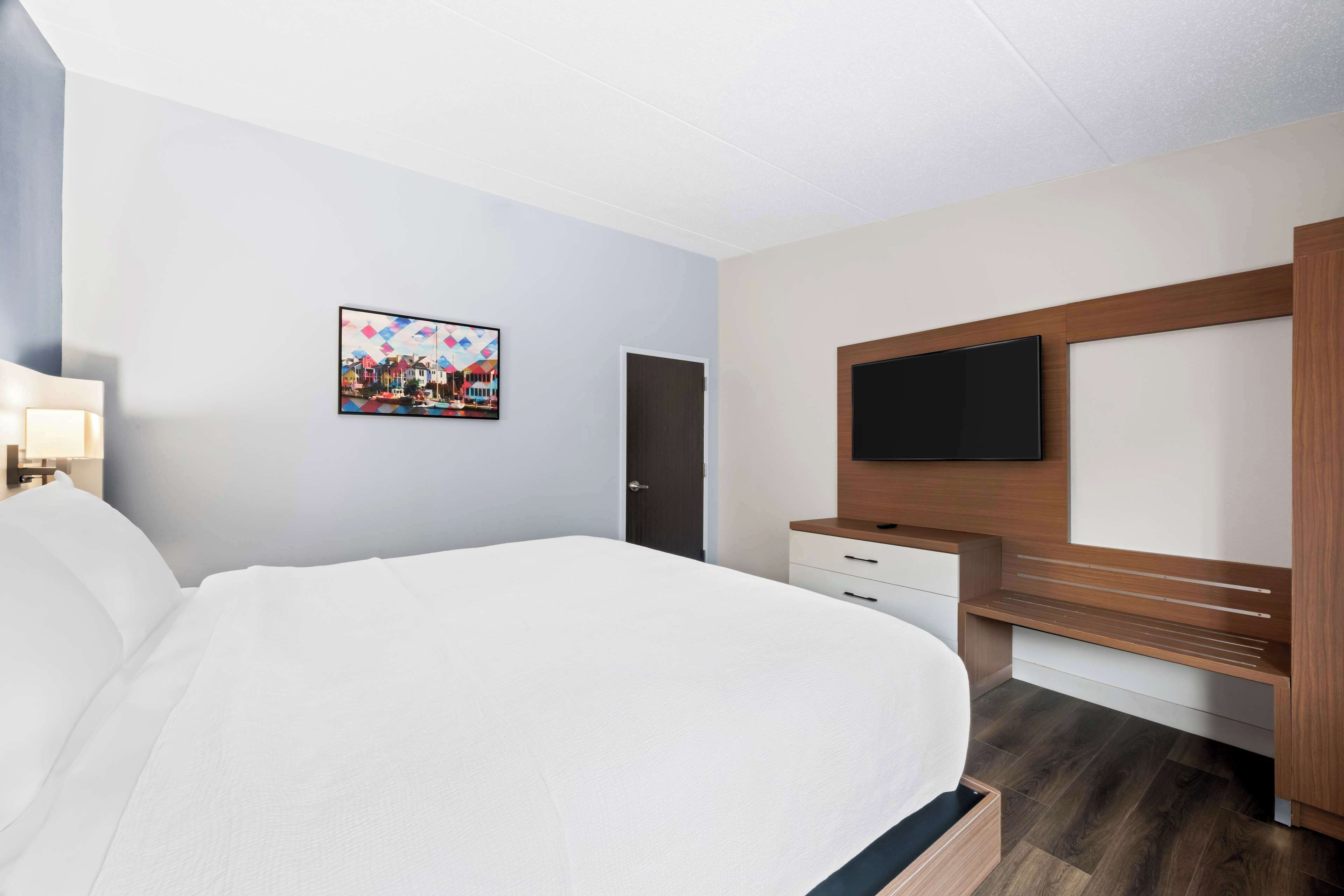 Quarto de hotel com uma cama grande, uma televisão de tela plana na parede, uma mesinha de cabeceira e uma porta.