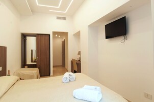 Quarto de hotel com uma cama, uma televisão presa à parede, uma mesa com uma cadeira e uma porta que dá para outro quarto.