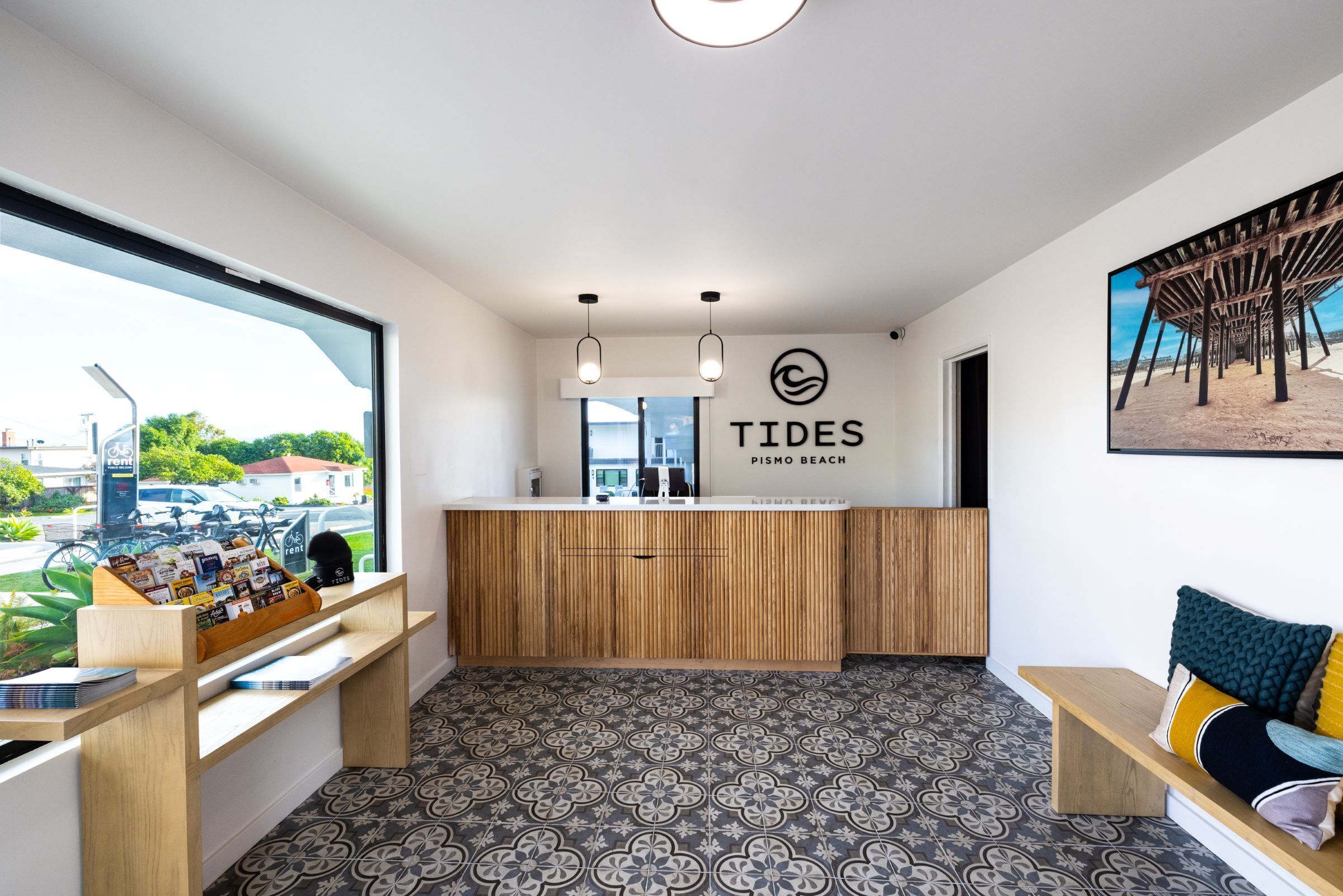 Uma área de recepção com balcão de madeira, uma placa na parede com a inscrição "TIDES PISMO BEACH" e piso estampado.