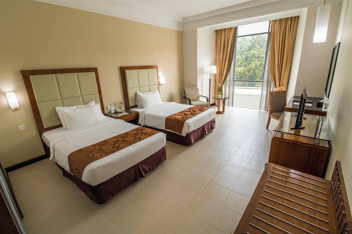 Quarto de hotel com duas camas, uma escrivaninha, uma cadeira, televisão e janela com cortinas.