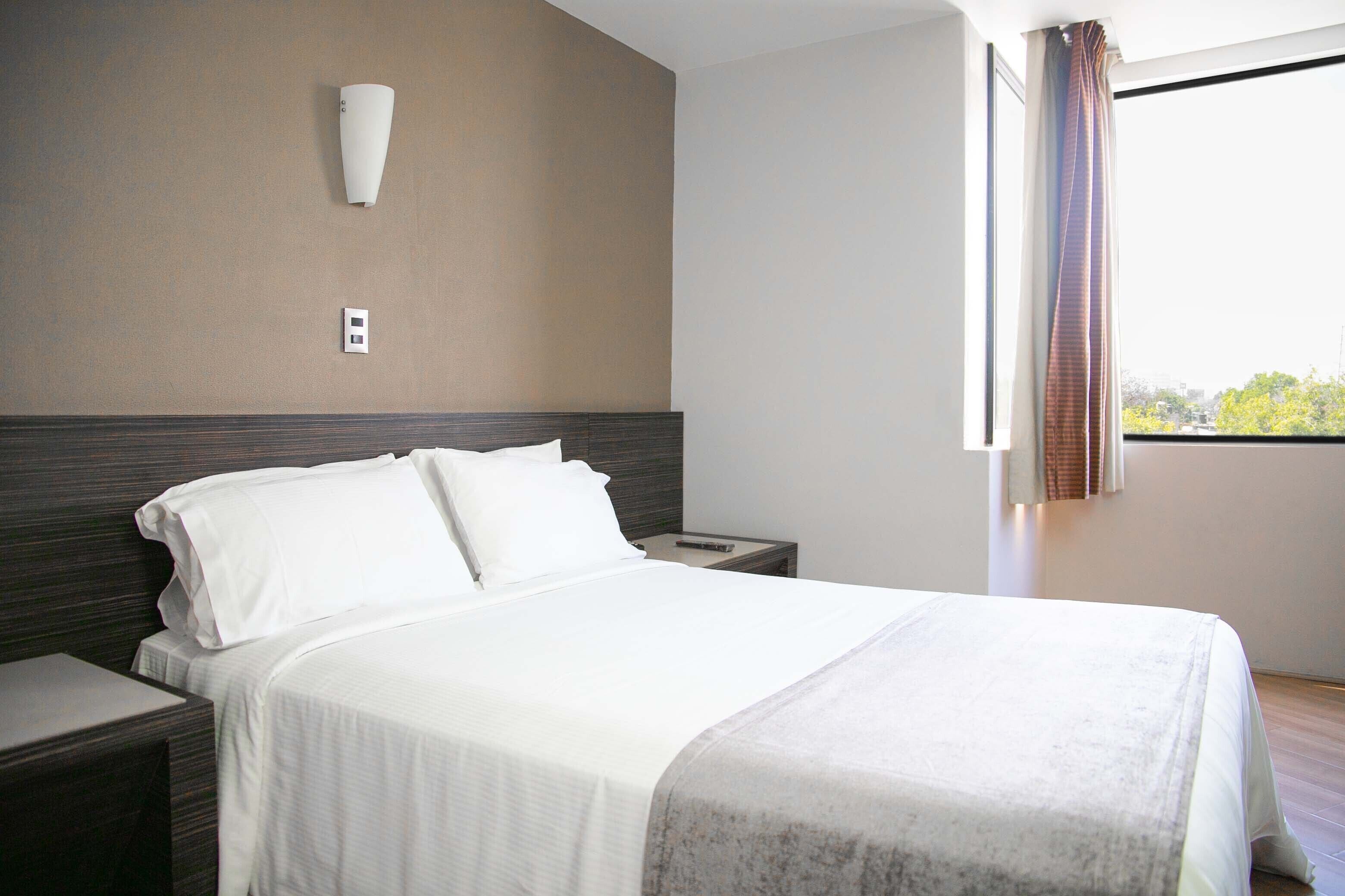 Quarto de hotel com cama, mesas de cabeceira, luminária na parede e janela com cortinas.