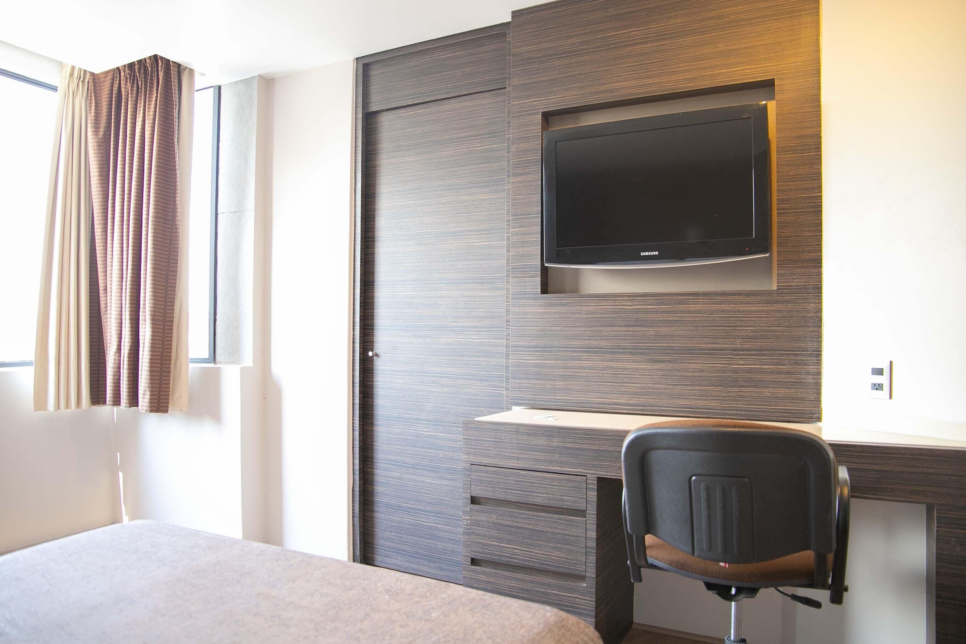 Quarto de hotel com uma cama, escrivaninha, cadeira, televisão fixada na parede e uma janela com cortinas.