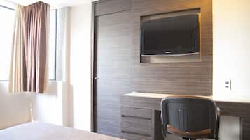 Quarto de hotel com uma cama, escrivaninha, cadeira, televisão fixada na parede e uma janela com cortinas.