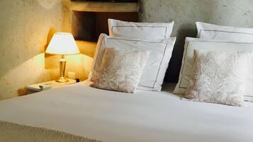 Cama bem arrumada com lençóis brancos e almofadas decorativas, uma luminária de cabeceira e uma estante com livros e um objeto decorativo.