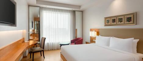 Quarto de hotel com uma cama grande, uma escrivaninha, uma cadeira, uma televisão e uma janela com cortinas.