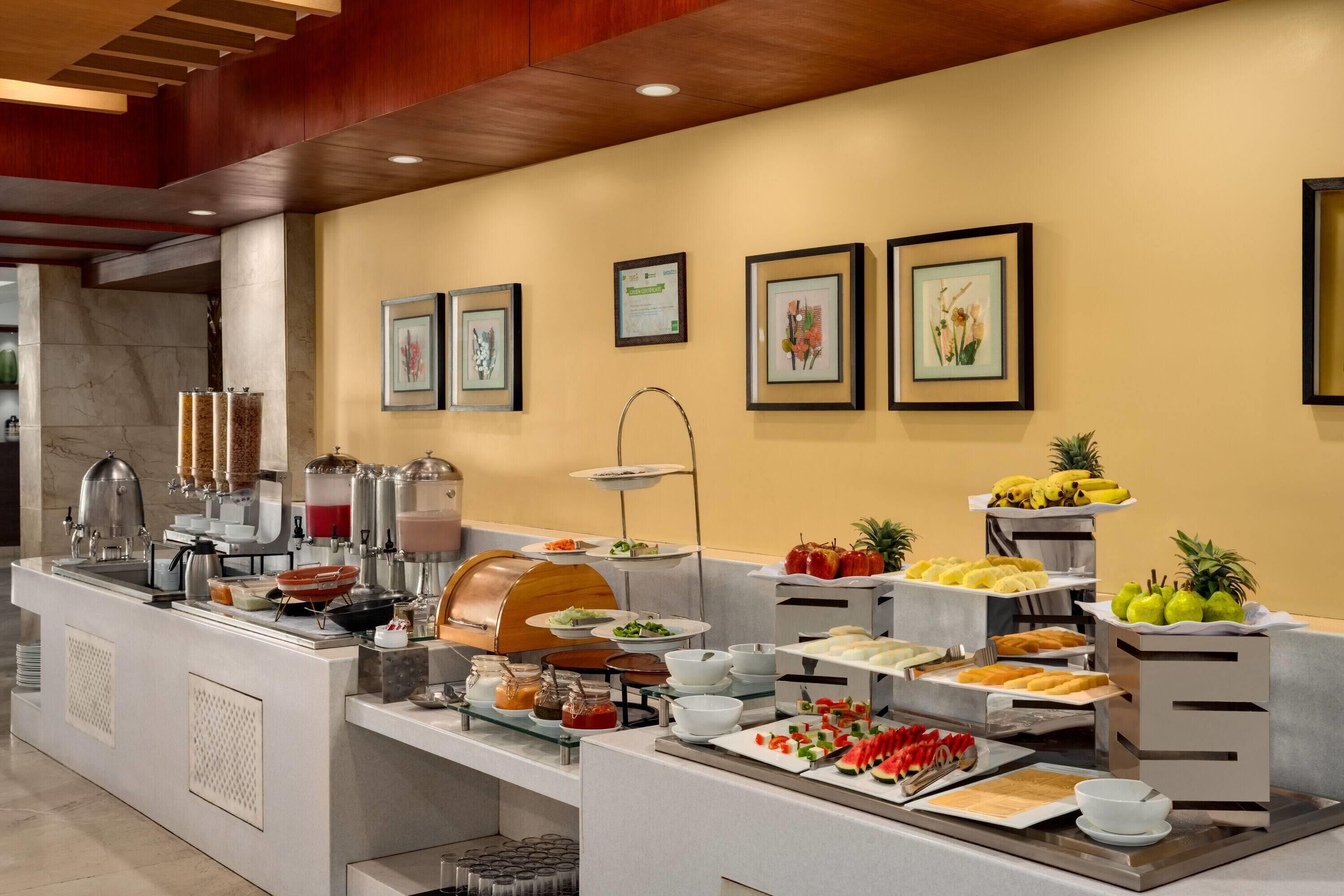 Disposição de buffet com diversos alimentos e bebidas, incluindo frutas, doces e um espaço para café.