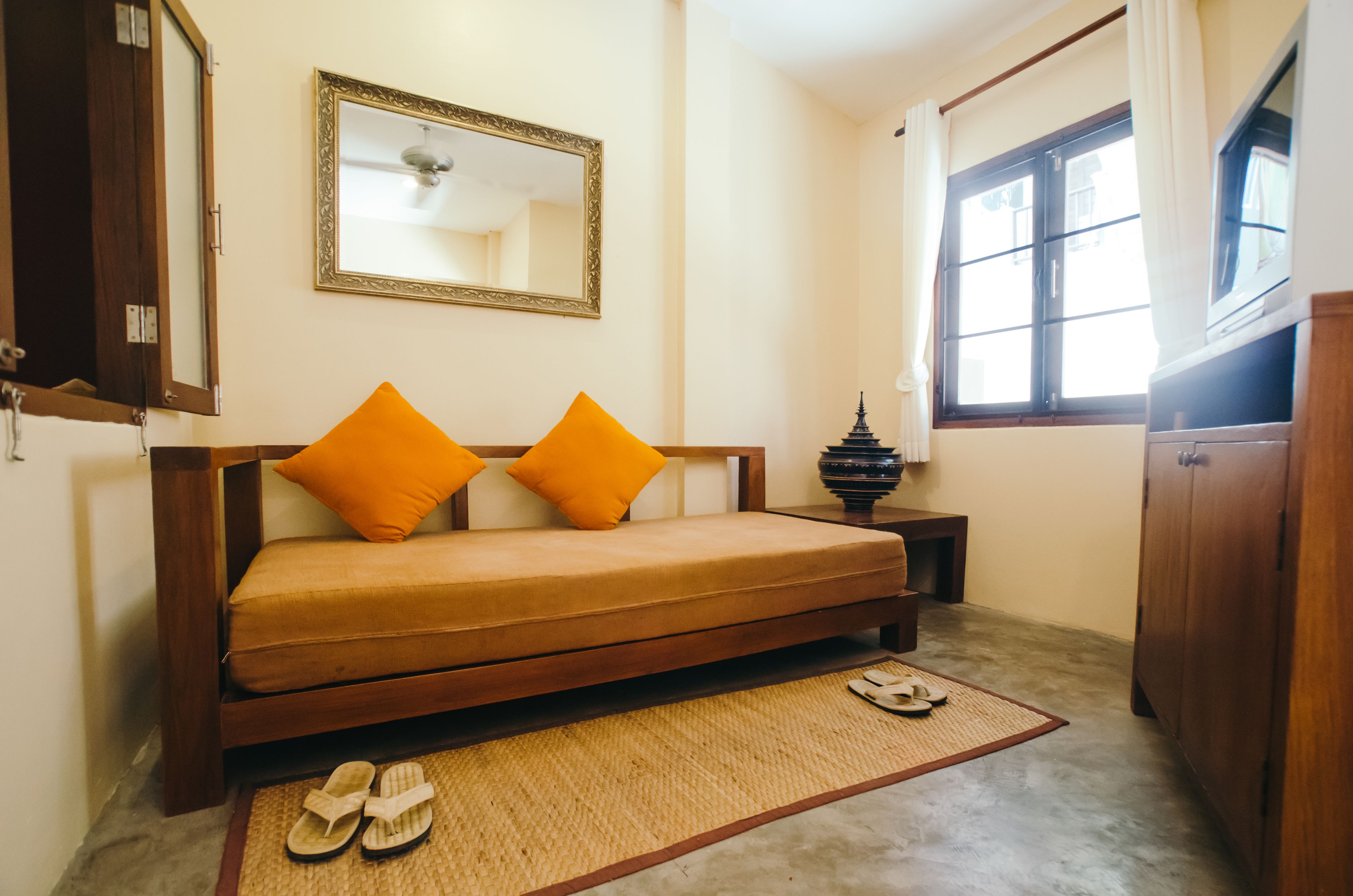 Quarto com cama de madeira, duas almofadas laranja, um espelho, uma janela com cortinas e uma pequena mesa com abajur.
