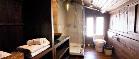 Casa de madeira com banheiro, incluindo box de vidro, um banco de madeira com toalhas, pia e vaso sanitário.