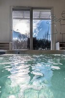 Piscina com água cristalina, vista para árvores e montanhas por uma porta de vidro, e uma escultura de metal na parede.