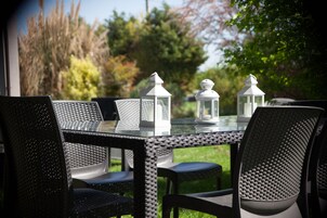 Uma mesa com tampo de vidro, cadeiras de vime, lanternas brancas e vista para árvores e gramado.