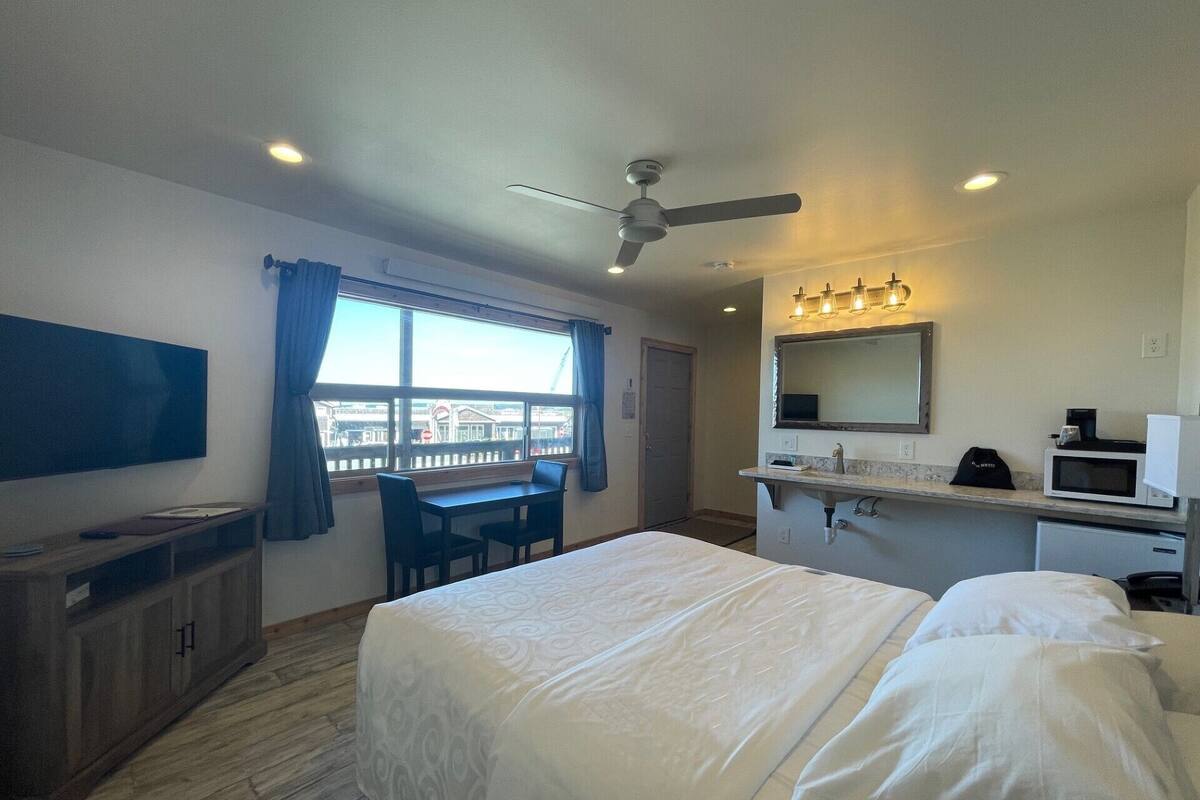 Quarto de hotel com cama, escrivaninha e cadeira, janela com vista, ventilador de teto e cozinha compacta com micro-ondas.