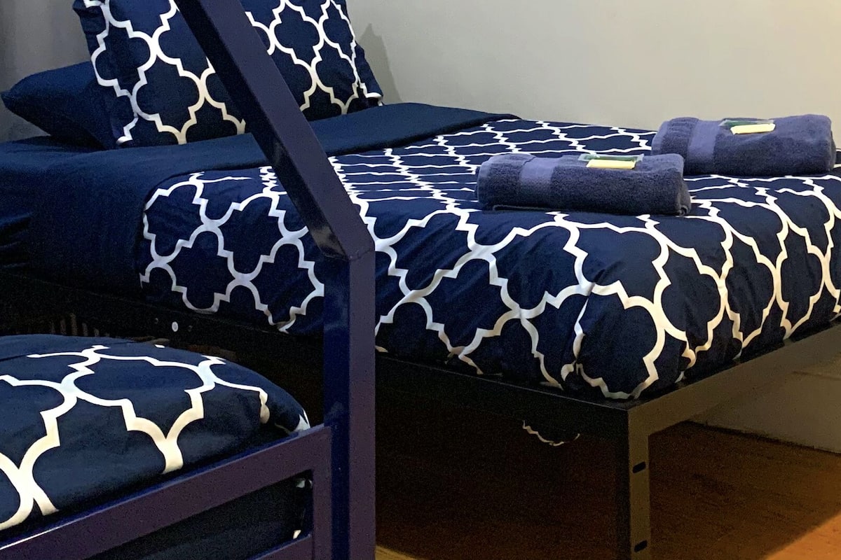 Um quarto com uma cama beliche e uma cama de solteiro, ambas com roupa de cama azul e branca estampada. Há uma planta em vaso sobre uma mesa redonda pequena ao lado da cama.