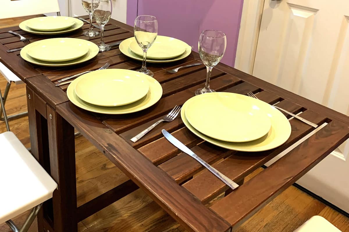 Uma mesa de jantar posta com pratos verdes, talheres e copos de vinho, com uma cadeira branca e um armário branco ao fundo.