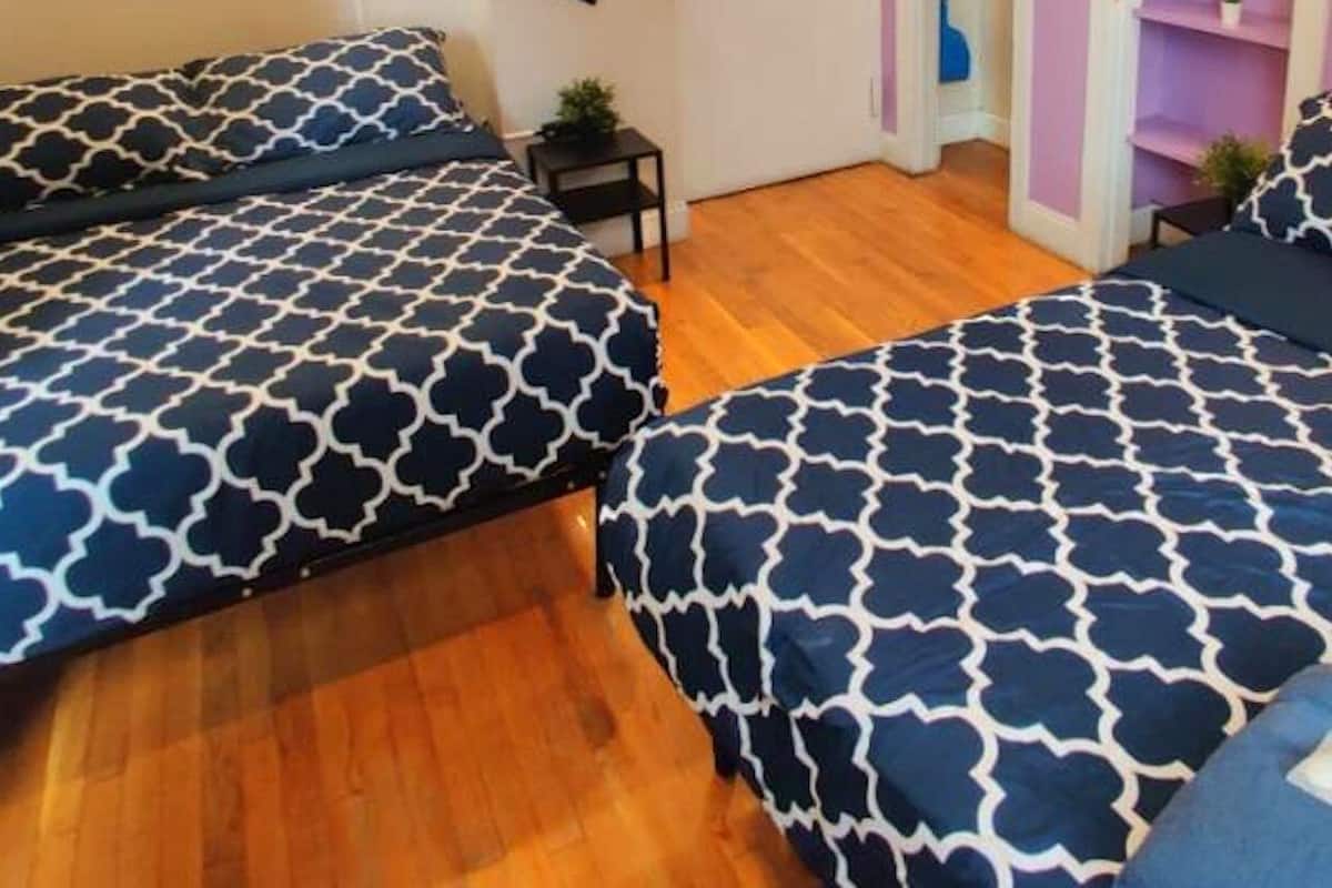 Quarto com três camas, cada uma com colcha geométrica azul e branca. Há uma mesinha com uma planta e uma estante com objetos decorativos.