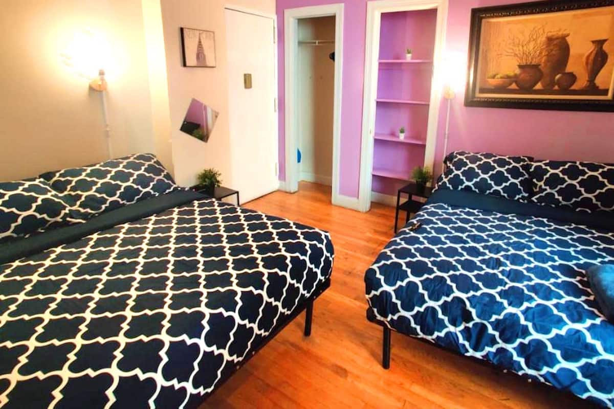 Quarto com duas camas, piso de madeira, uma porta, uma prateleira com plantas e um quadro na parede.