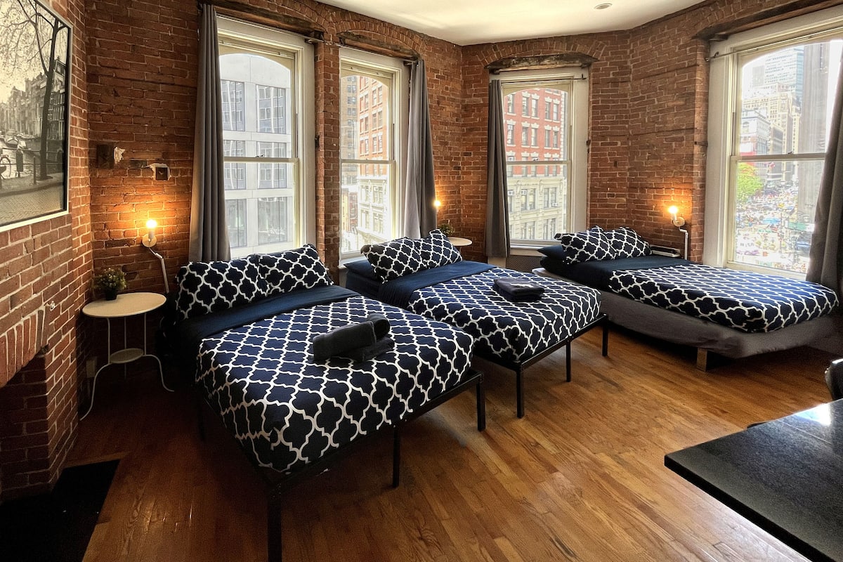 Quarto com paredes de tijolo, grandes janelas e vista da cidade. Há três camas com roupa de cama estampada e uma pequena mesa com abajur.