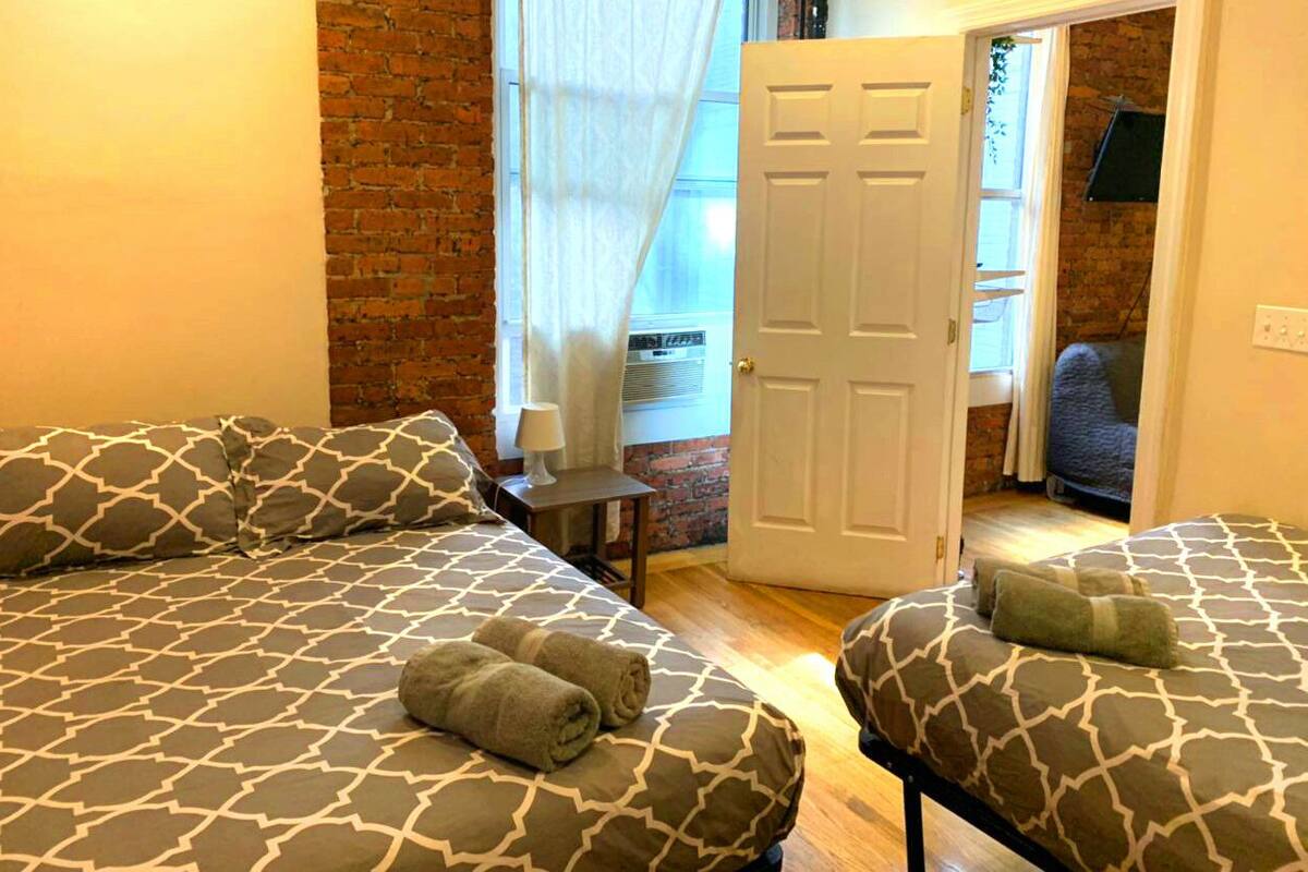 Quarto com duas camas, cada uma com roupa de cama estampada e toalhas dobradas. Há uma porta que dá para outro quarto, uma janela com cortinas e uma pequena mesa com abajur.