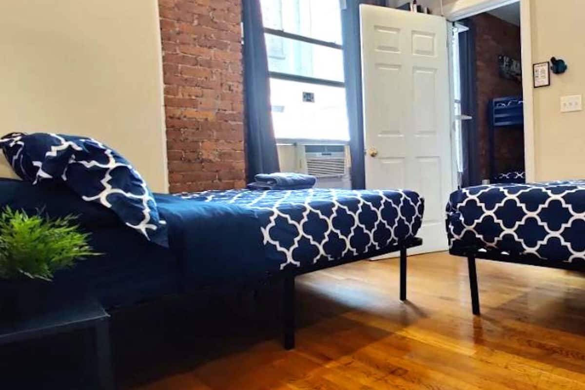 Quarto com parede de tijolos, assoalho de madeira, cama com roupa de cama estampada, televisão montada na parede e porta que dá para outro quarto.