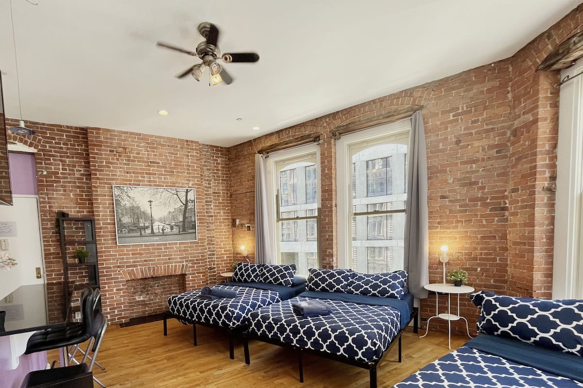 Quarto com parede de tijolos, ventilador de teto, cama com roupa de cama estampada, mesa pequena com abajur e janela com cortinas.