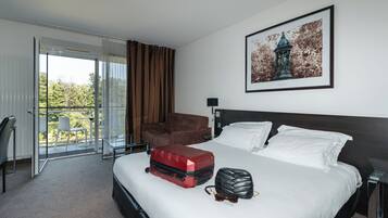Quarto de hotel com uma cama, uma mala vermelha, uma bolsa preta e um par de óculos de sol sobre a cama. Há uma cadeira marrom e uma mesa de vidro com uma cadeira. É visível uma varanda com porta de vidro e cortinas.