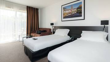Quarto de hotel com duas camas, uma cadeira, uma escrivaninha e uma janela grande com cortinas.