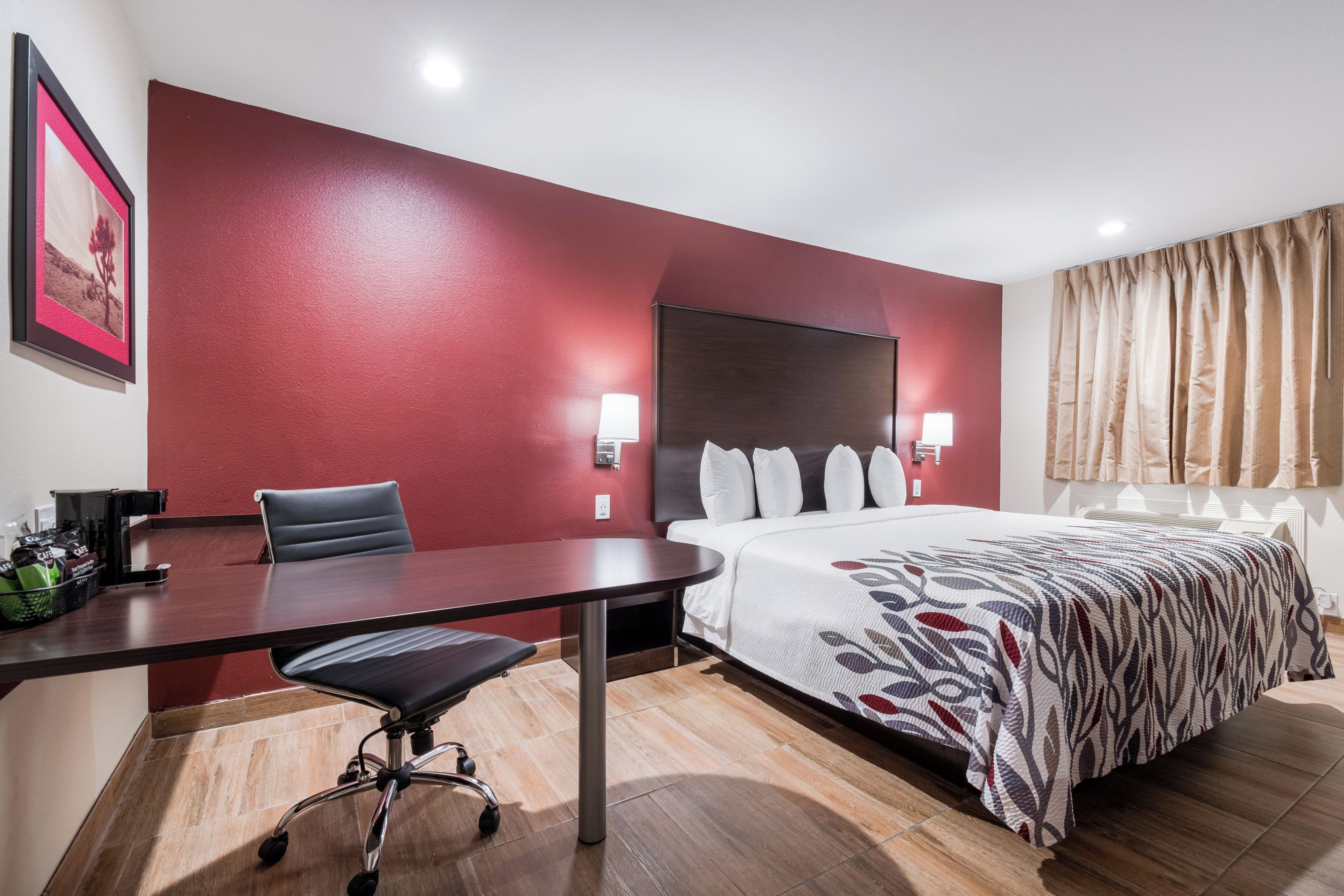 Quarto de hotel com cama, escrivaninha, cadeira, luminária e janela com cortinas.