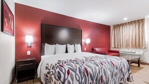 Quarto de hotel com uma cama grande, uma cadeira vermelha, uma escrivaninha e uma janela com cortinas.