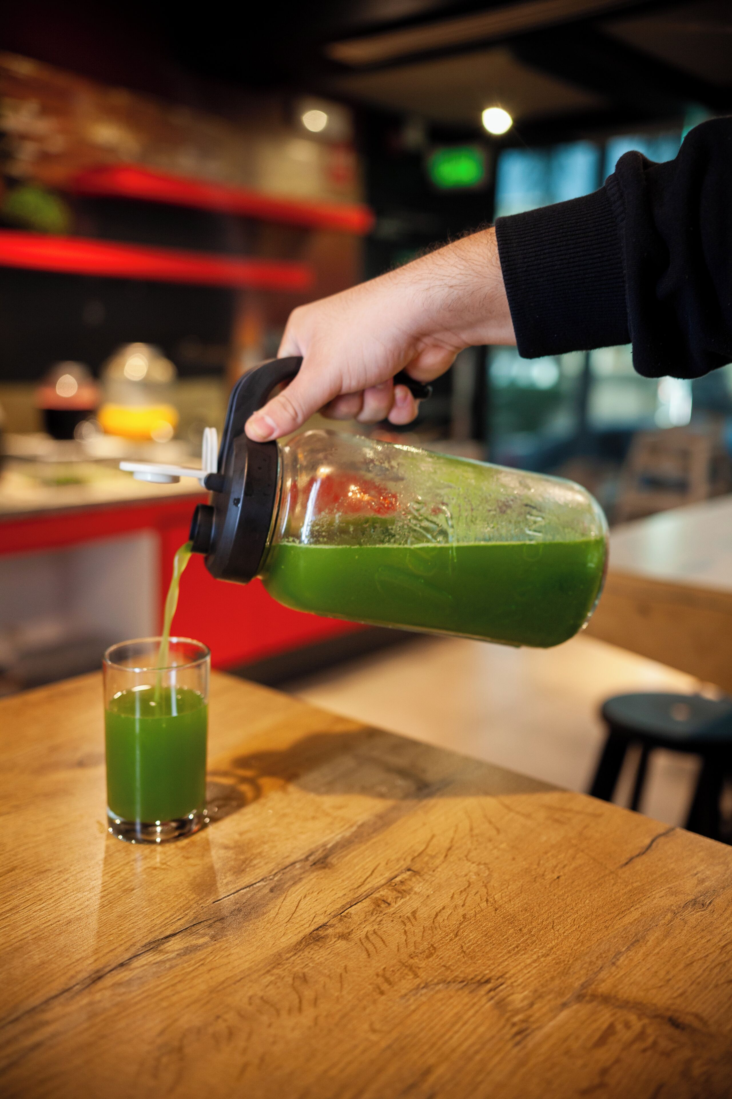 Uma mão despejando um líquido verde de um coqueteleira para dentro de um copo sobre uma mesa de madeira.