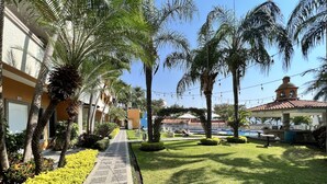 Uma passagem bordeada por palmeiras leva a uma área de piscina com assentos externos e uma estrutura coberta.