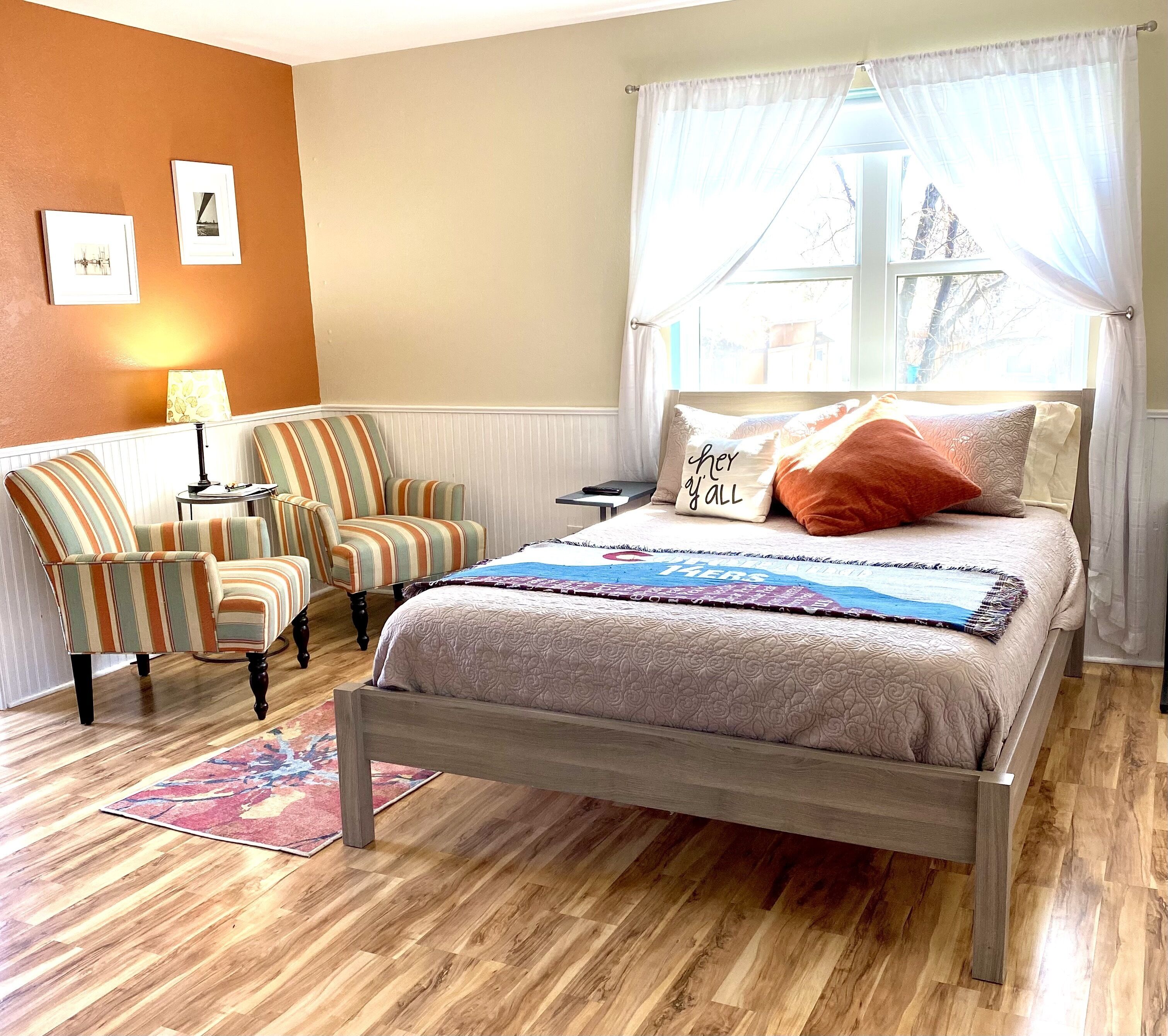 Quarto com cama, duas poltronas listradas, mesa de cabeceira, luminária e janela com cortinas.