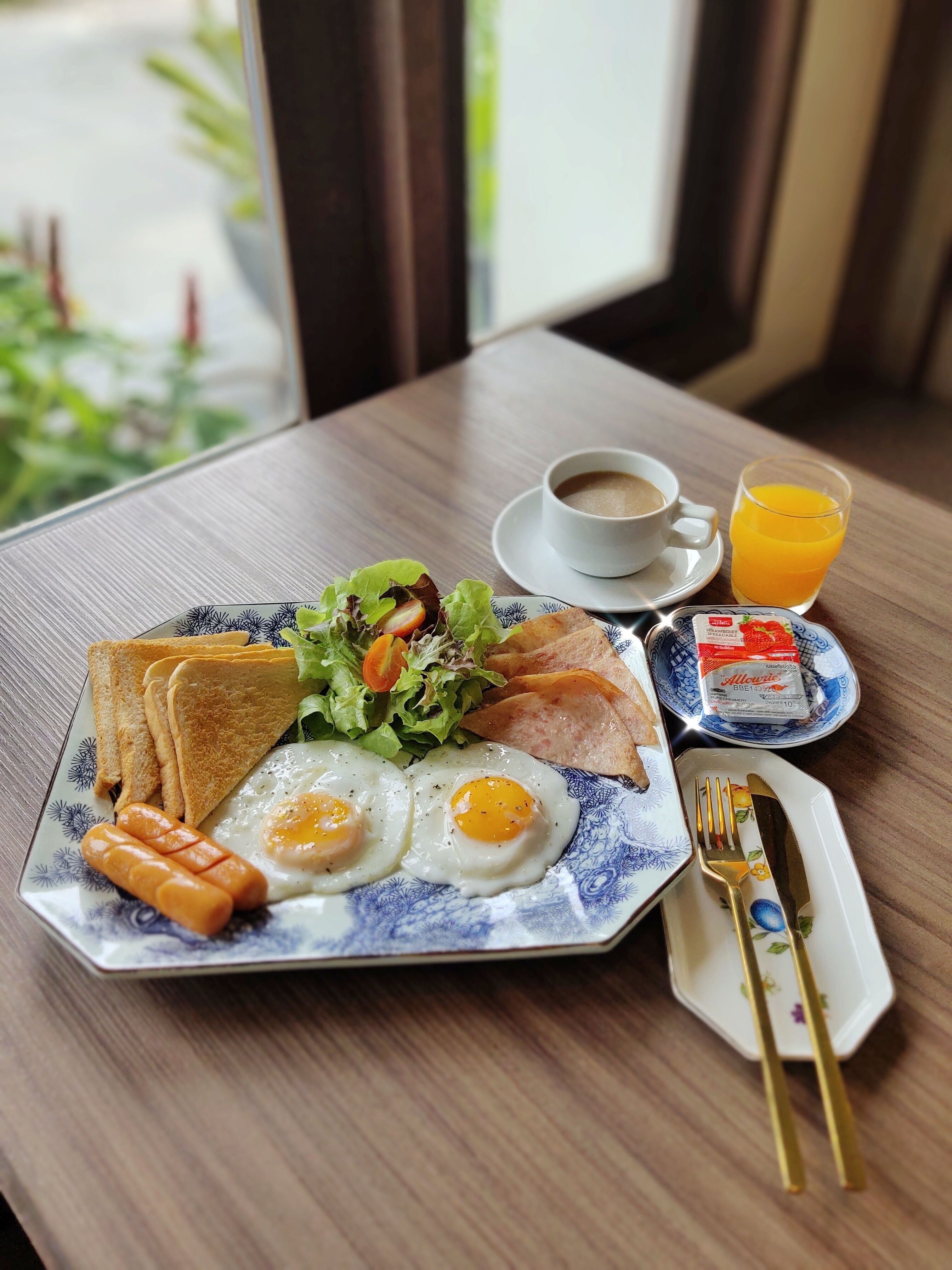 Prato de pequeno-almoço com torradas, ovos, salsichas e uma salada, uma chávena de café e um copo de sumo de laranja sobre uma mesa de madeira.