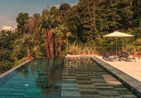 Piscina com água cristalina, rodeada por vegetação exuberante e árvores. Há espreguiçadeiras e um guarda-sol.