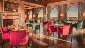 Restaurante com cadeiras vermelhas, lareira e amplas janelas arqueadas com vista para a cidade.