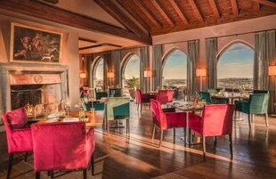 Restaurante com cadeiras vermelhas, lareira e amplas janelas arqueadas com vista para a cidade.